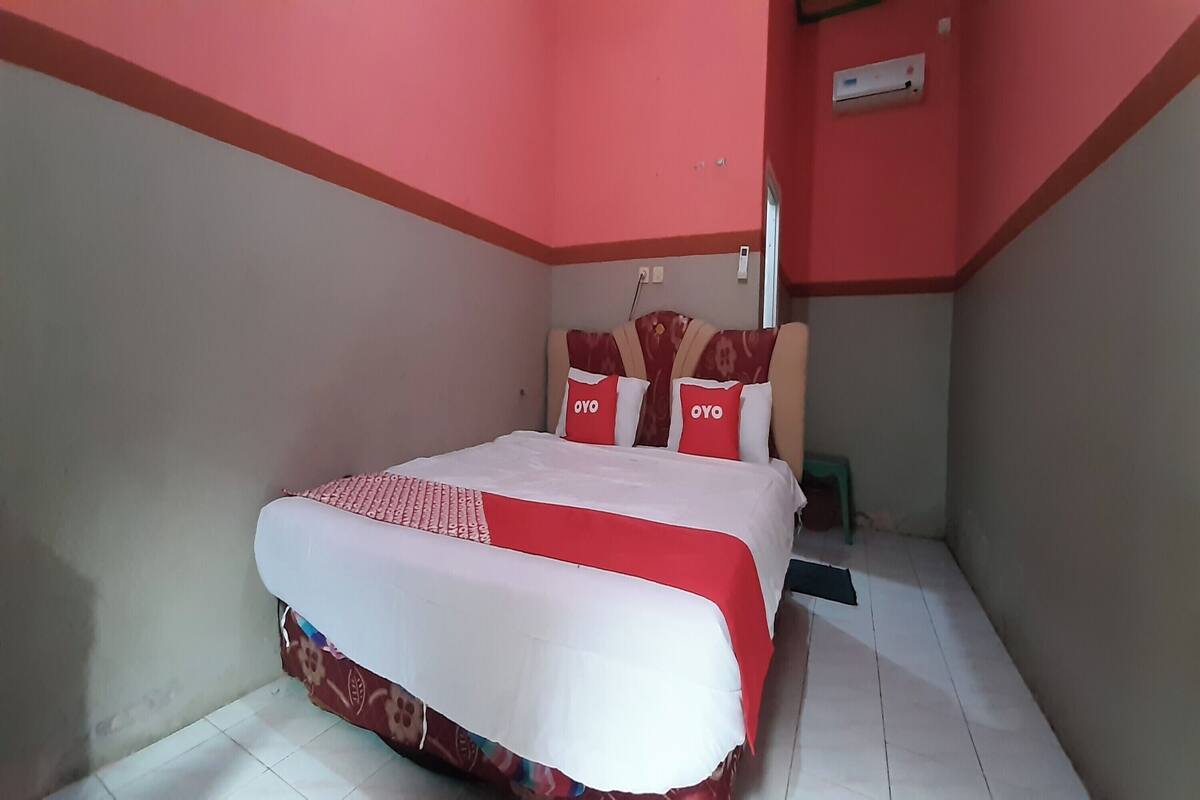 Quarto pequeno e estreito com uma cama, ar-condicionado fixado na parede e um banquinho.