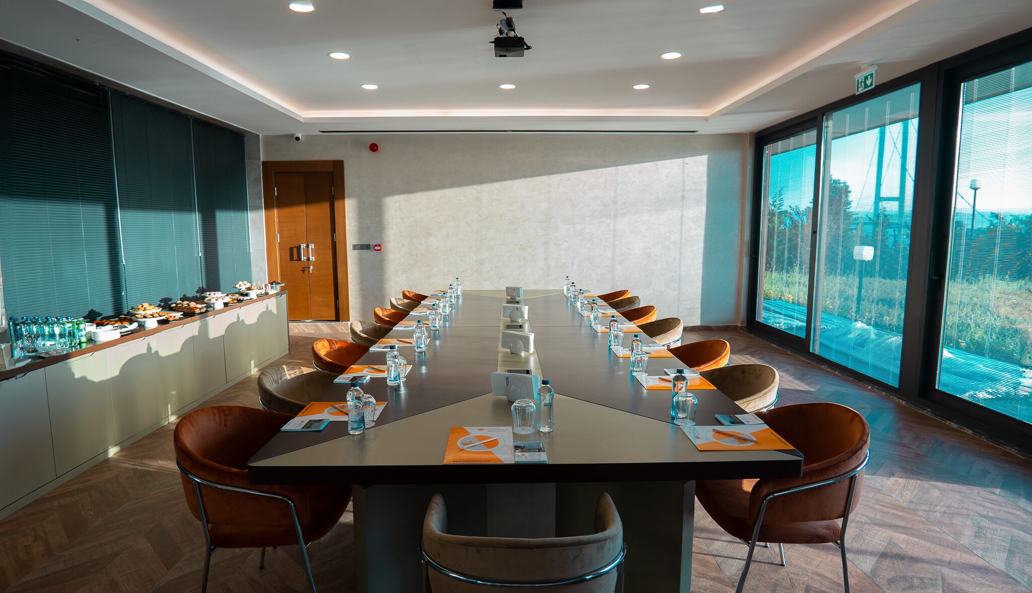 Sala de conferências com uma mesa comprida, cadeiras, garrafas de água e um buffet à esquerda.