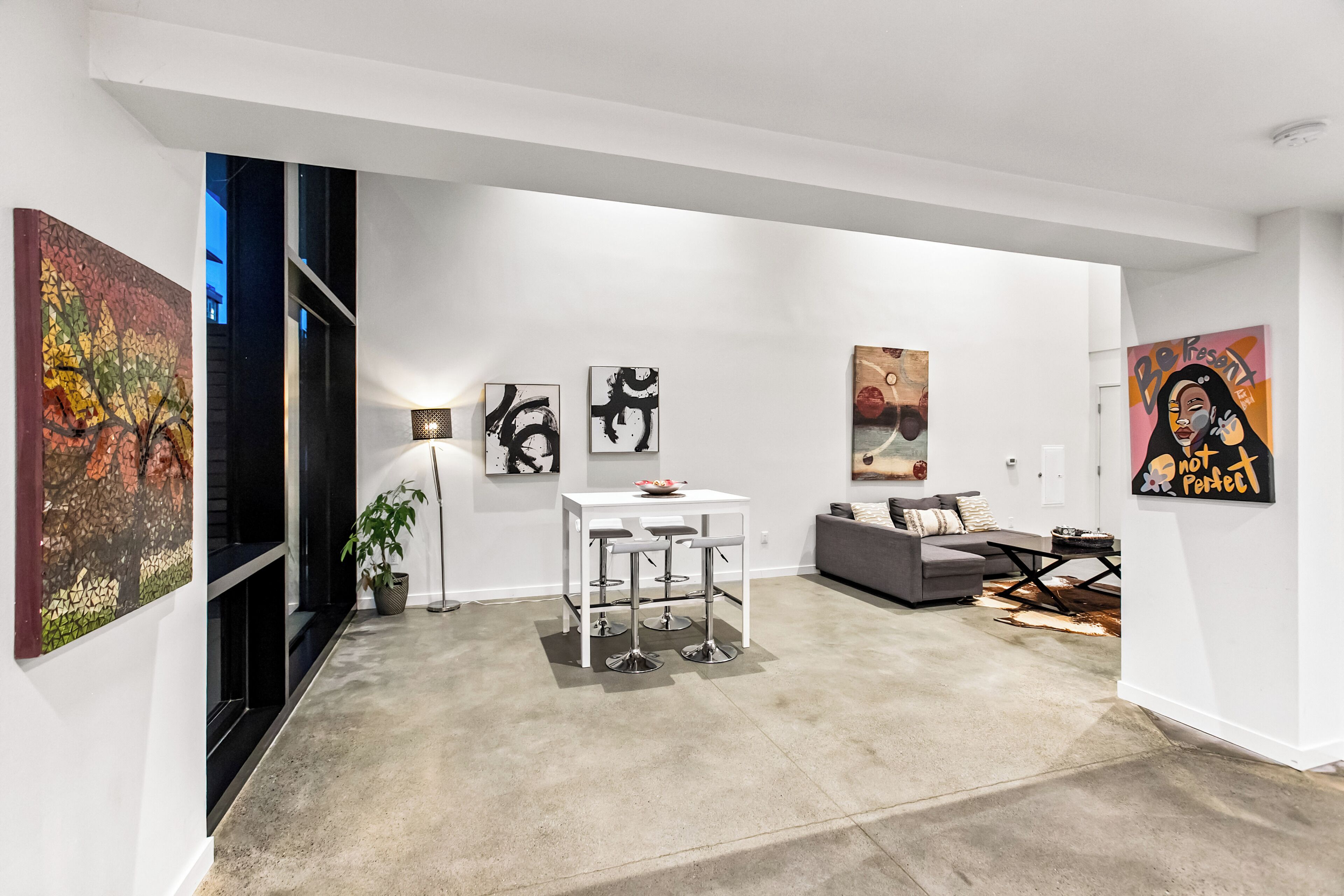 Uma galeria de arte com paredes brancas e chão de concreto, exibindo diversas obras de arte e uma pequena mesa branca com bancos.