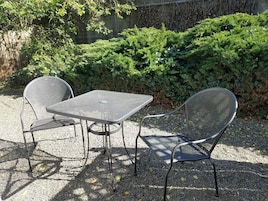 Mesa de metal e duas cadeiras em ambiente externo, com chão de cascalho e folhagem verde ao fundo.