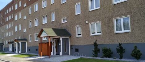 Um hotel de vários andares com fachada de tijolos, uma entrada pequena com toldo verde e um gramado bem cuidado.