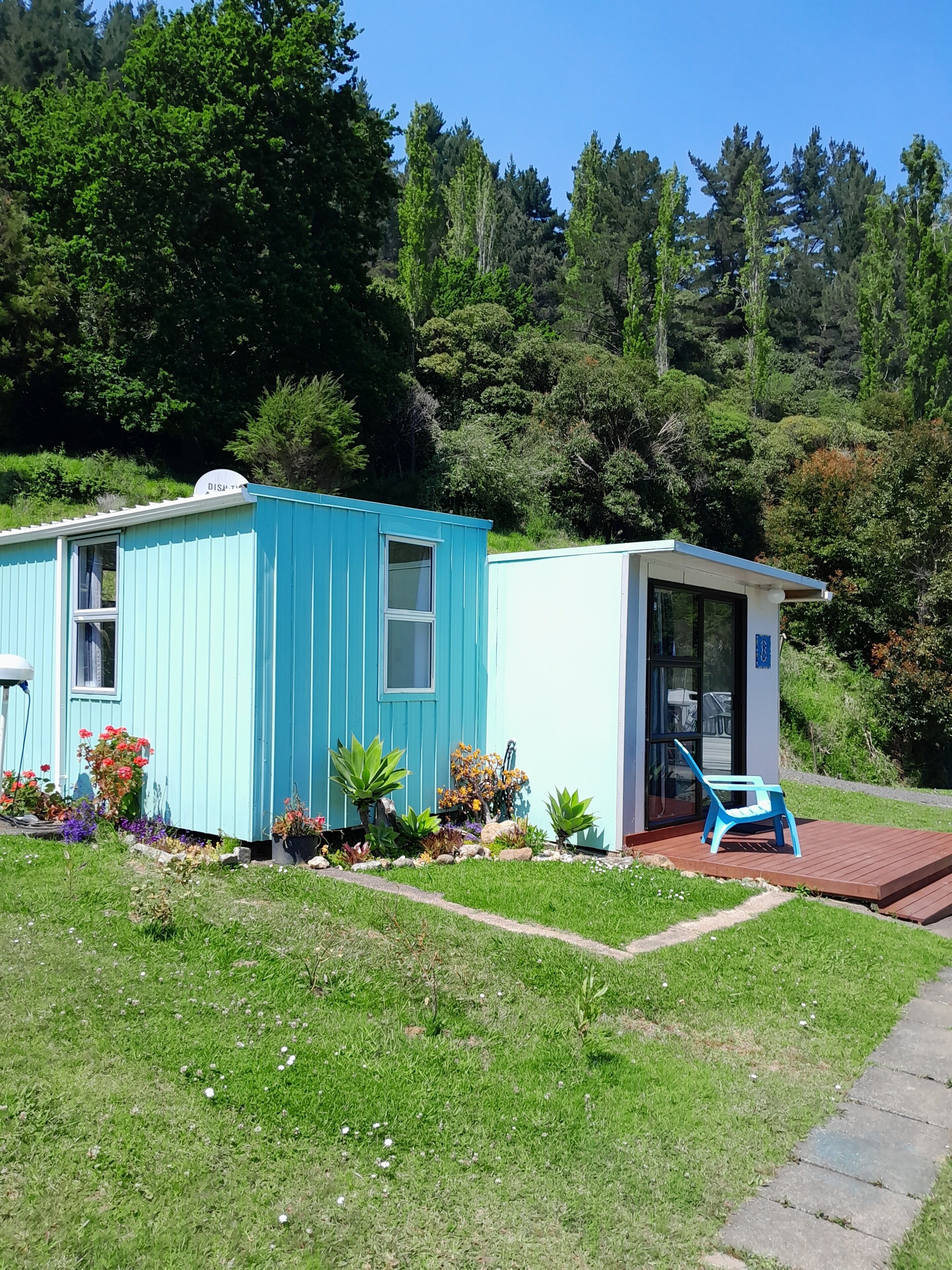 Um pequeno edifício térreo com fachada azul clara, um pequeno alpendre com uma cadeira azul e um jardim com diversas plantas e flores.