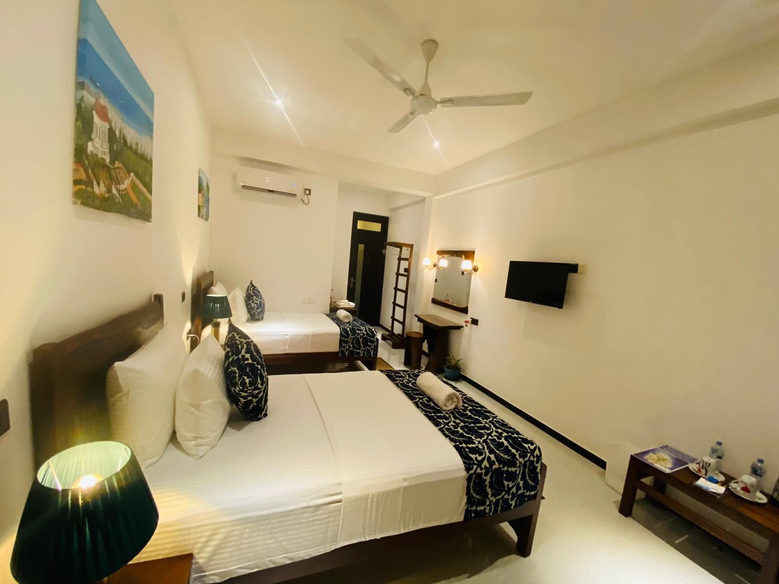 Quarto de hotel com duas camas, um ventilador de teto, televisão, banheiro com espelho e um quadro na parede.