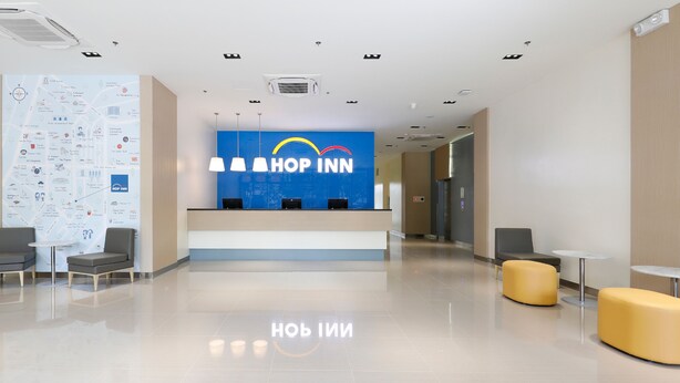 Uma área de recepção de hotel moderna, com uma parede azul exibindo o logotipo 'HOP INN', um balcão de recepção e disposição de assentos incluindo cadeiras e pufes.