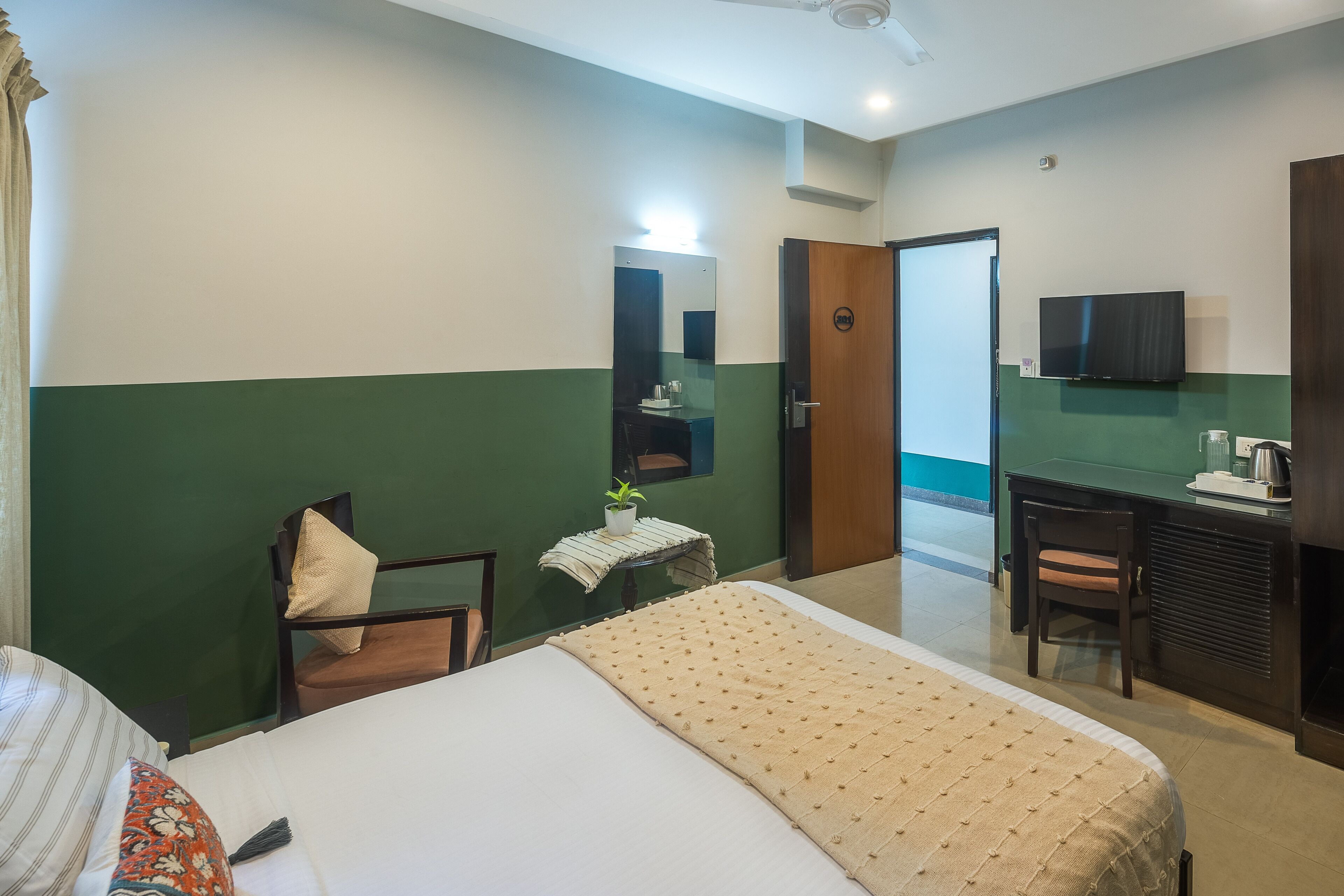 Quarto de hotel com cama, escrivaninha, cadeira, pequena mesa com uma planta, espelho e televisão.