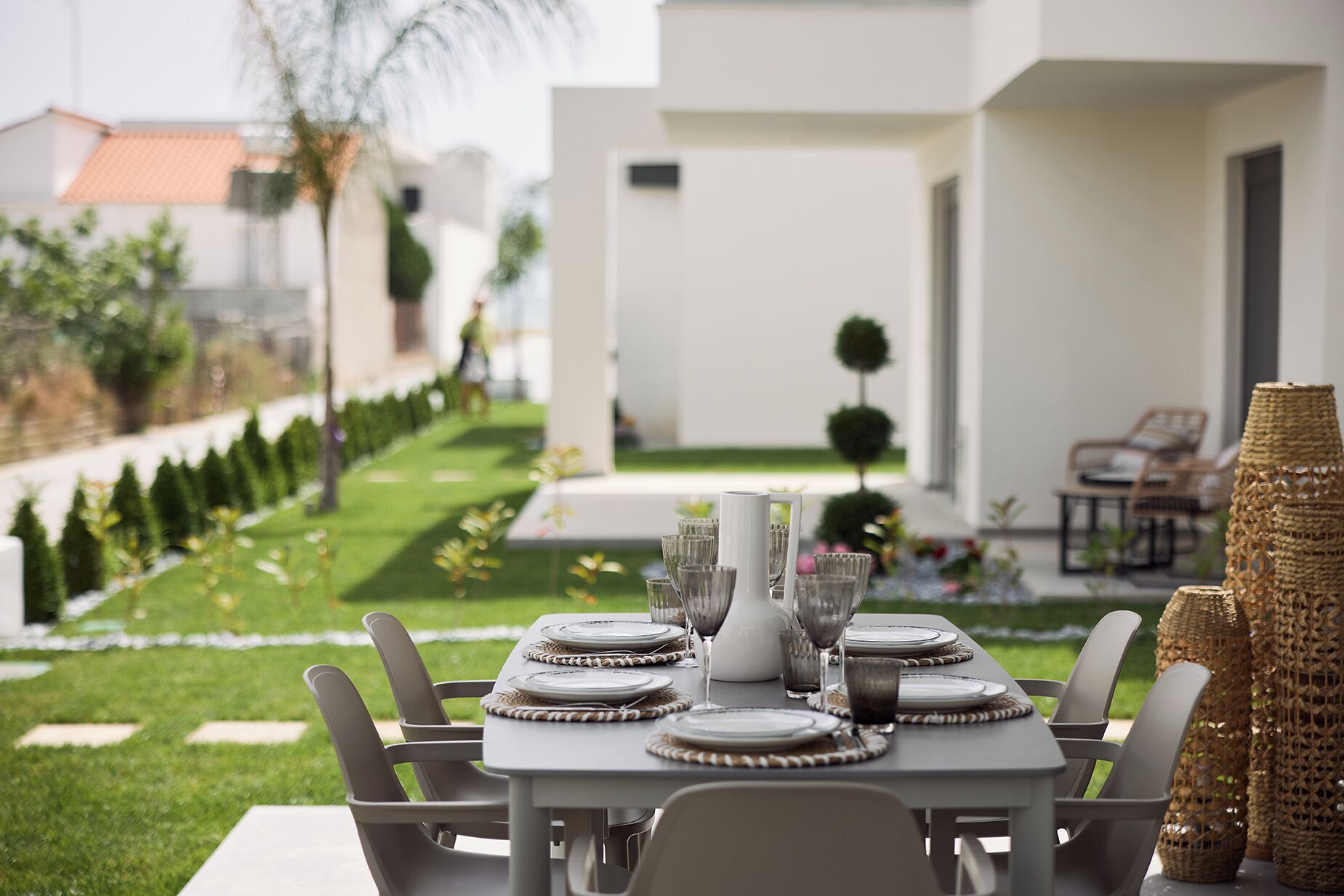 Uma área de refeições ao ar livre com uma mesa posta para seis pessoas, rodeada por cadeiras, em uma casa moderna com um jardim bem cuidado.