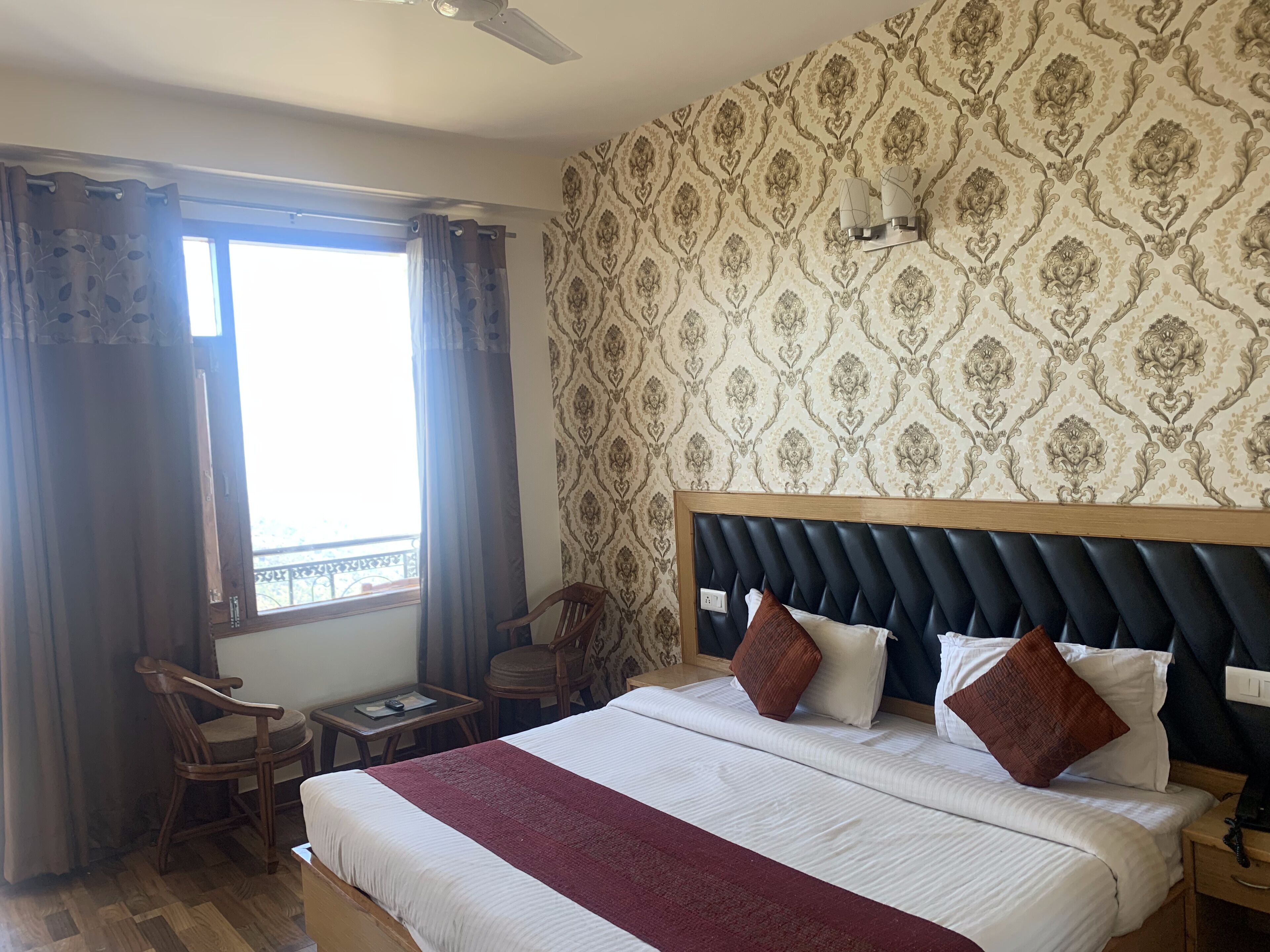 Quarto de hotel com uma cama grande, cabeceira de madeira, mesa de cabeceira, uma cadeira, uma mesinha e uma janela com cortinas.