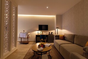 Uma sala de estar moderna com um sofá, uma mesa de centro com frutas e um livro, uma mesa de jantar com toalha branca e uma TV fixada na parede.