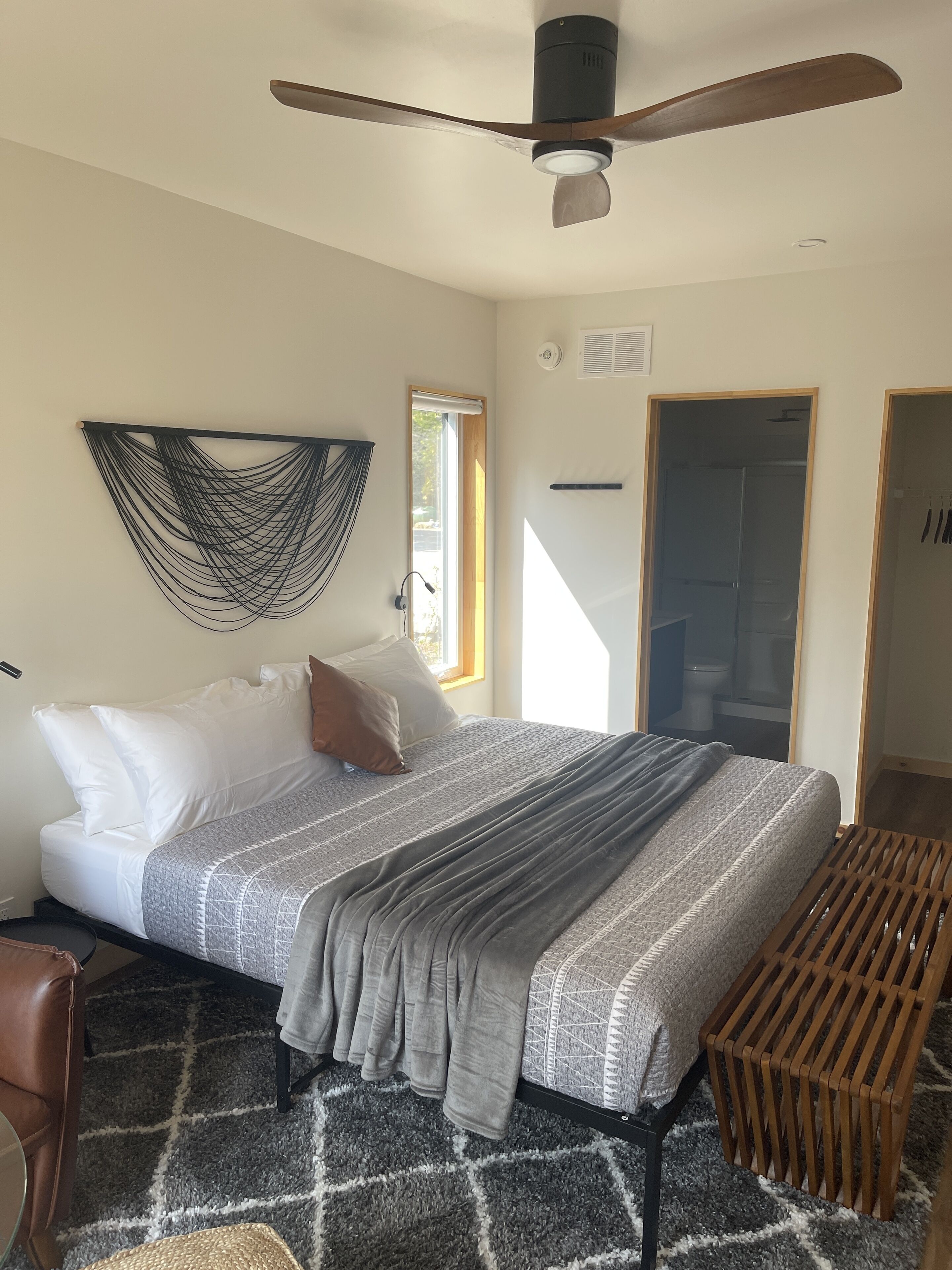 Quarto com cama, ventoinha no teto, luminária na parede, janela e porta que dá para um banheiro.