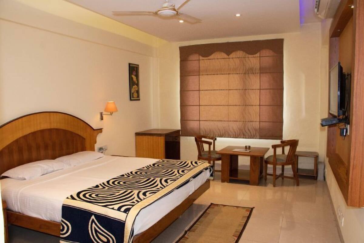 Quarto com cama, escrivaninha, cadeira, luminária, ventoinha de teto e uma grande janela com detalhe em madeira.