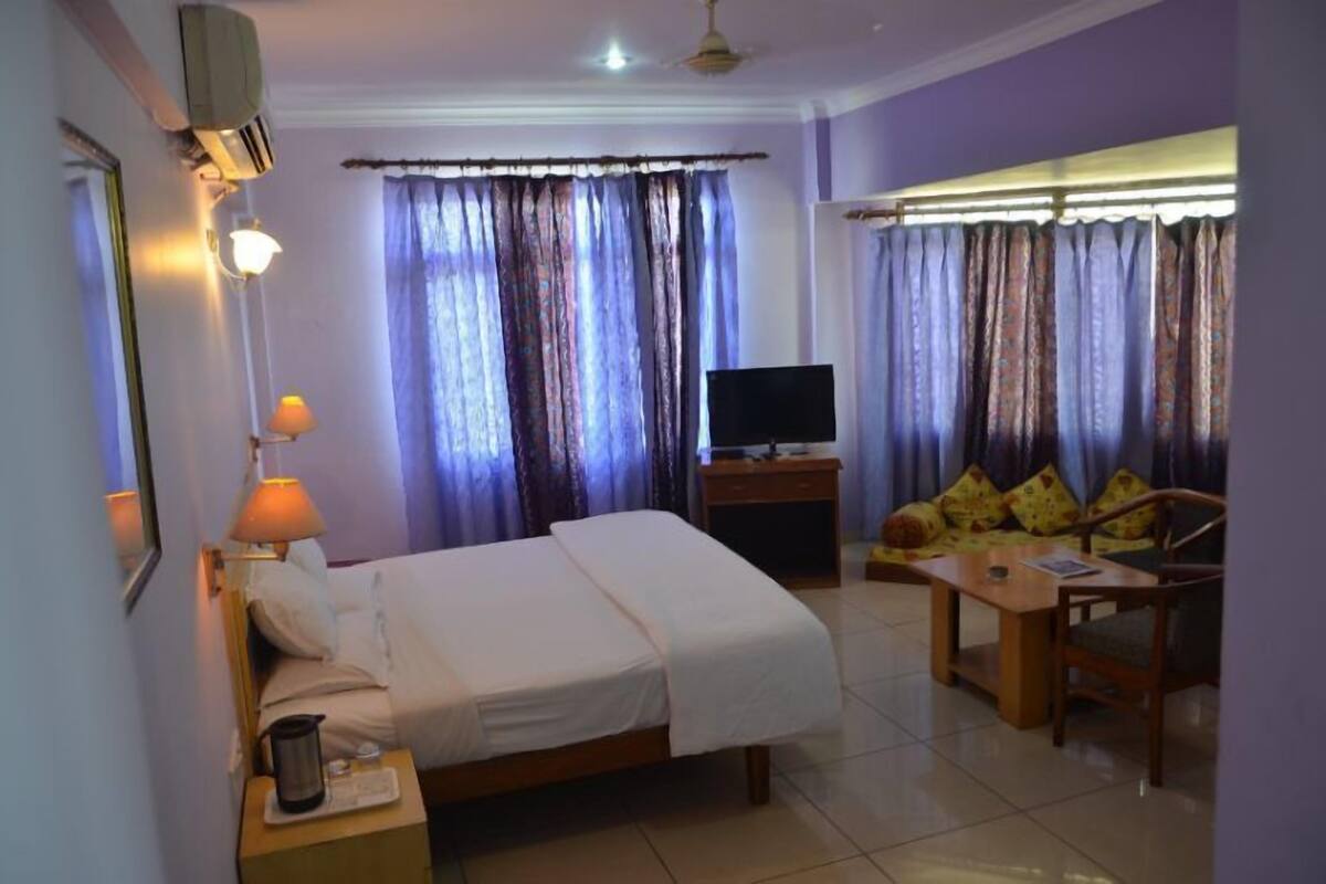 Quarto de hotel com cama, televisão, escrivaninha e cadeira, além de cortinas.