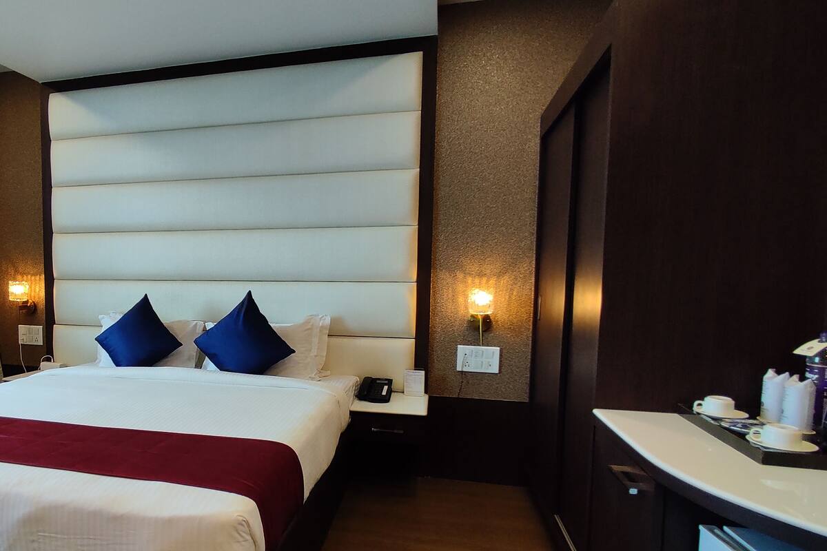 Quarto de hotel com uma cama grande, duas almofadas, uma mesa de cabeceira com telefone, uma mesinha com chávena e pires, e uma janela com cortinas.