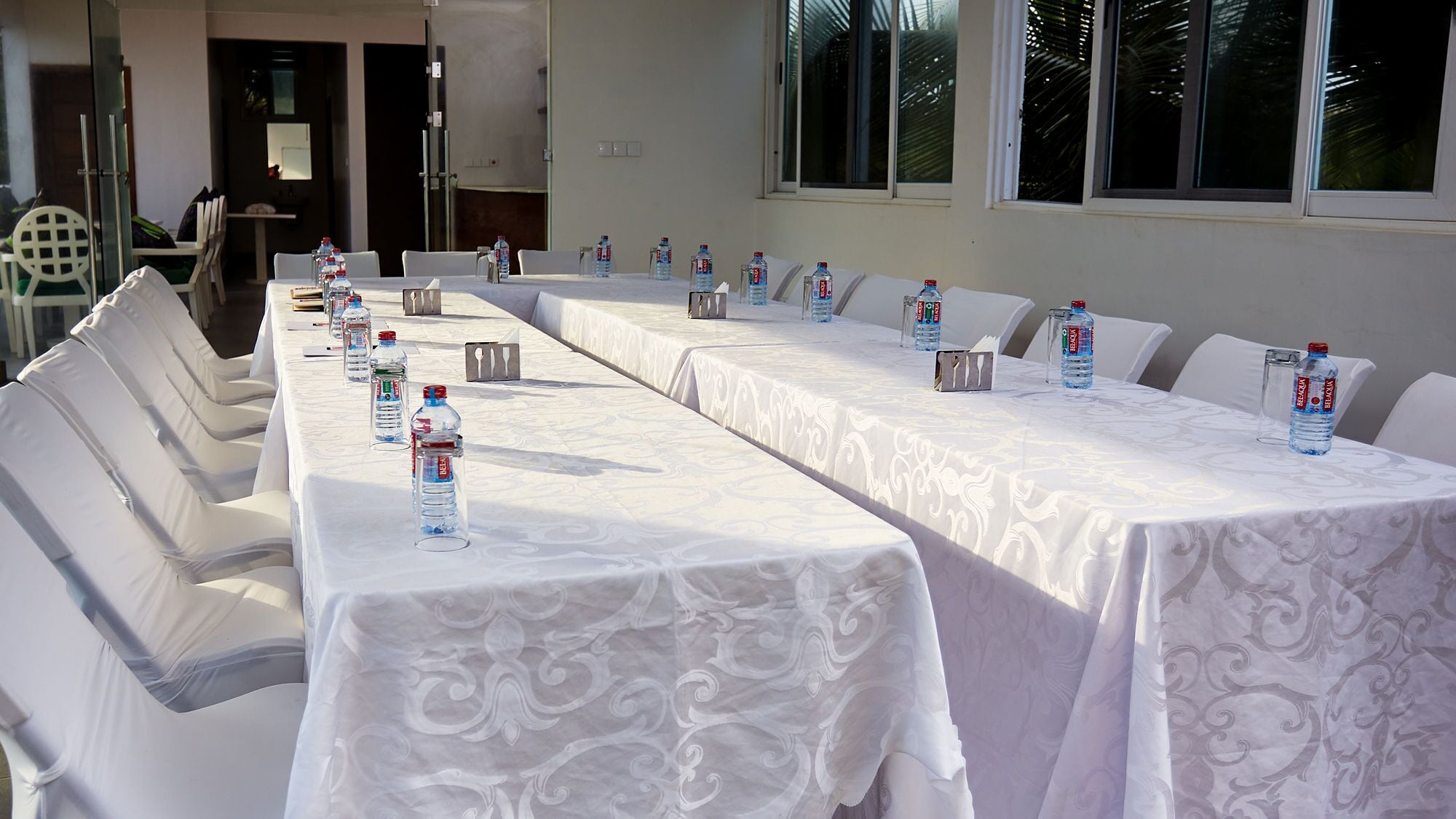 Uma mesa comprida com toalhas brancas e cadeiras, garrafas de água e um copo em cada mesa.