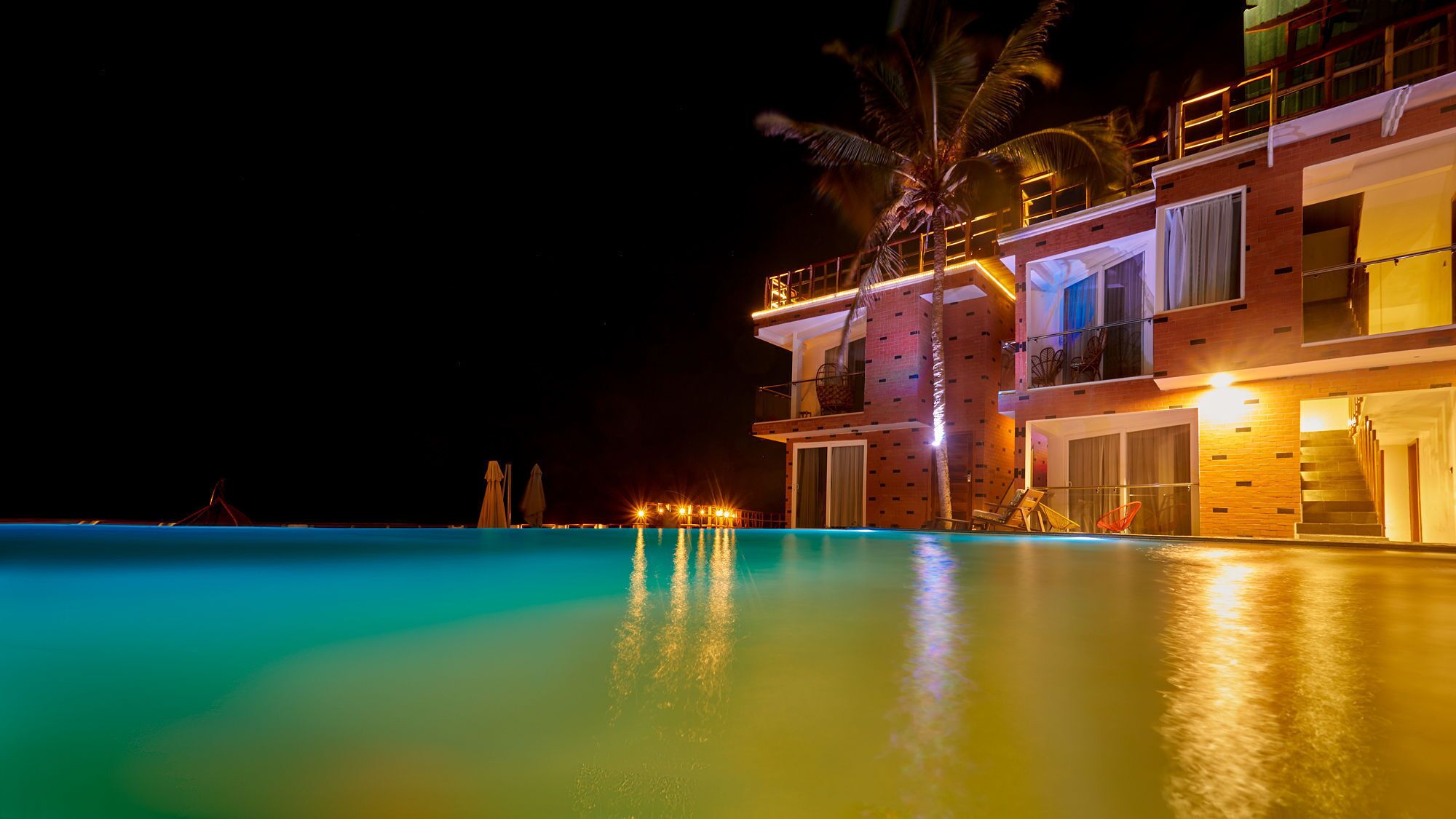 Vista noturna de um hotel com piscina, uma palmeira e uma varanda bem iluminada.