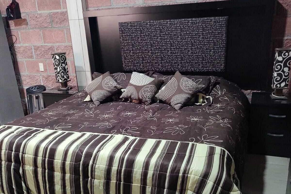 Cama com colcha listrada e várias almofadas, parede de tijolos ao fundo e uma pequena mesa de cabeceira com luminária.