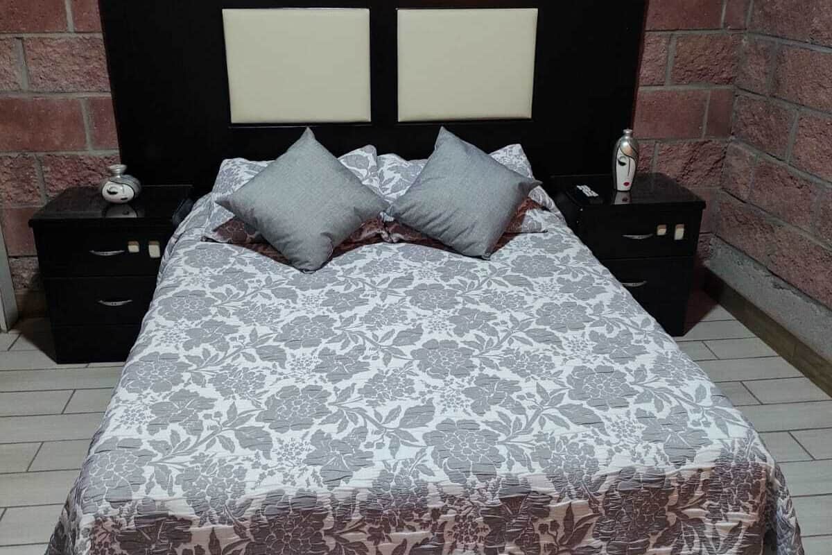 Uma cama com colcha estampada, duas almofadas e cabeceira com dois painéis. Há duas mesas de cabeceira, uma com um relógio e a outra com uma garrafa. O quarto tem uma parede de tijolos ao fundo.