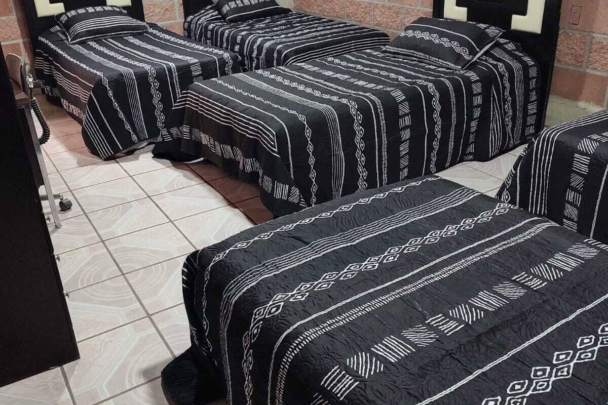 Quarto com quatro camas, cada uma com colcha e cabeceira estampada em preto e branco. O quarto possui piso de azulejos e paredes de tijolo.