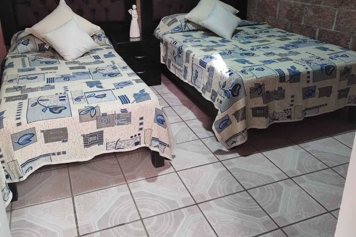 Duas camas de solteiro com roupa de cama estampada, um criado-mudo pequeno com uma figurinha, e um fundo de parede de tijolos.