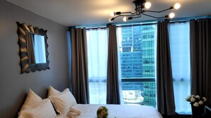 Quarto de hotel com uma cama grande, um candelabro moderno e vista para o horizonte urbano através de amplas janelas.