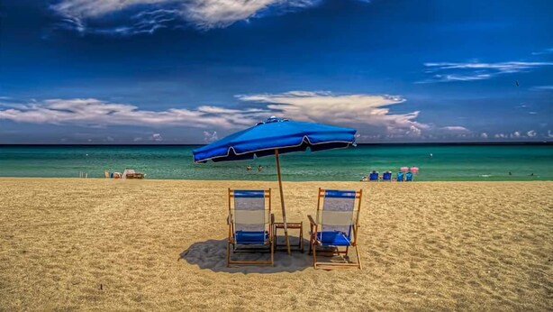 Uma praia com duas cadeiras de praia azuis e um guarda-sol azul sob um céu claro.