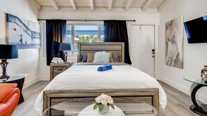 Quarto com uma cama grande, mesa de cabeceira, televisão, janela com cortinas e obras de arte nas paredes.