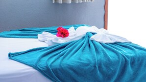 Quarto de hotel com cama coberta por toalha azul e lençol branco, uma flor rosa e uma janela com cortinas.