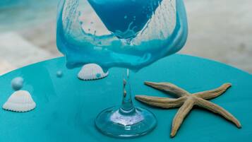 Um copo com líquido azul e uma cereja por cima, apoiado em uma superfície azul-turquesa com conchas e uma estrela-do-mar.