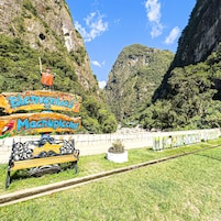 Uma placa de madeira com 'Bienvenue à Machupicchu' e uma ilustração de pássaro, um banco, um vaso de plantas e uma cerca branca com pinturas coloridas.