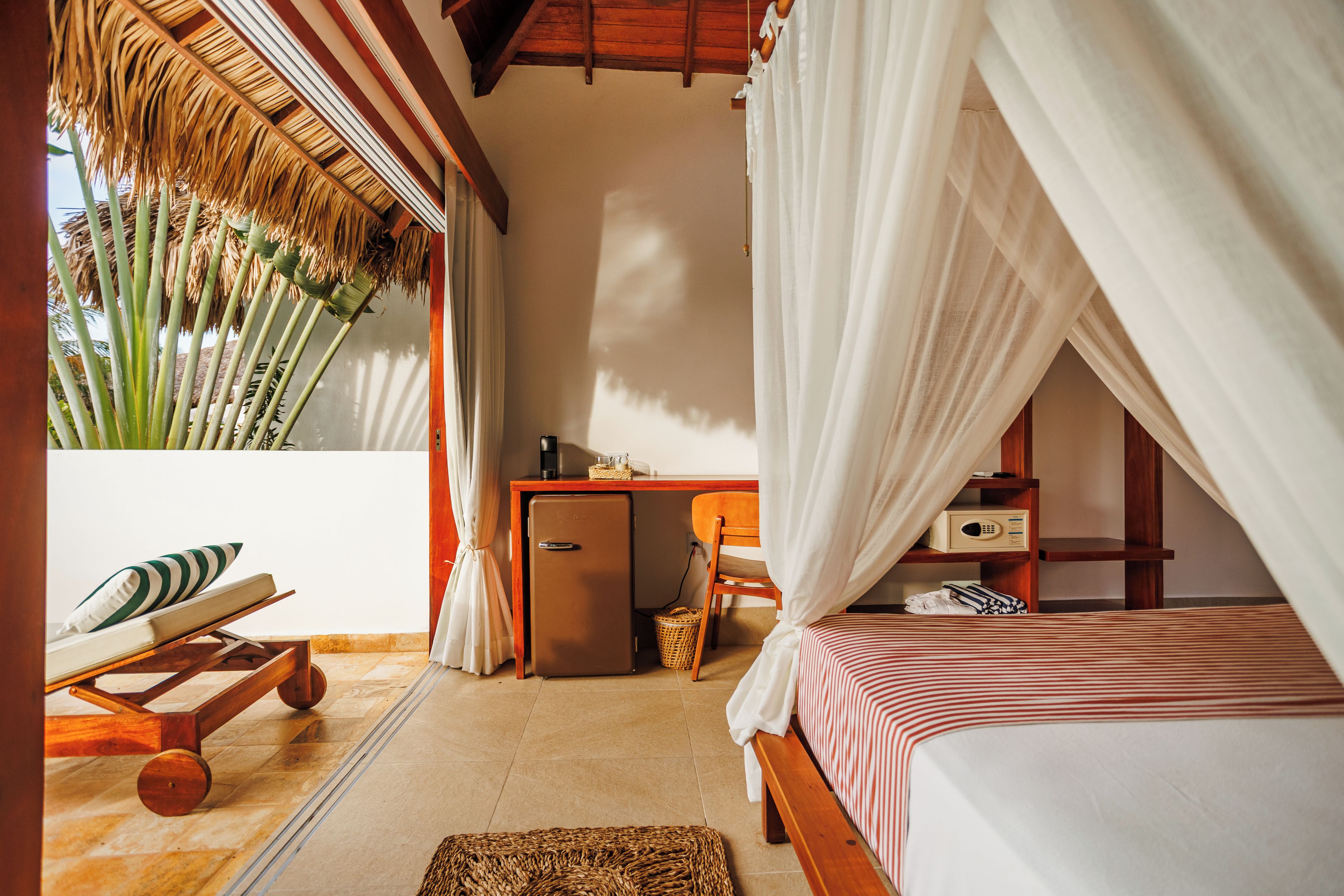 Quarto com cama, escrivaninha, cadeira, pequeno frigorífico e janela com vista para palmeiras.