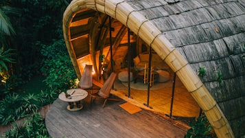 Estrutura de bambu com telhado curvo, deck de madeira e área de estar com mesa e cadeiras.
