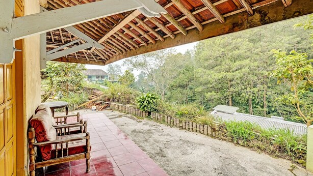 Uma área externa com mesa e banco de madeira, um alpendre coberto com piso de azulejos e vista para uma encosta arborizada.