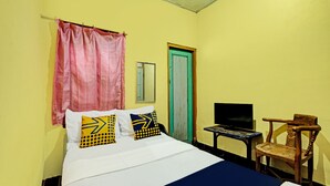 Cama com roupa de cama branca e almofadas decorativas, uma televisão sobre uma mesinha, uma cadeira de madeira e uma porta verde.