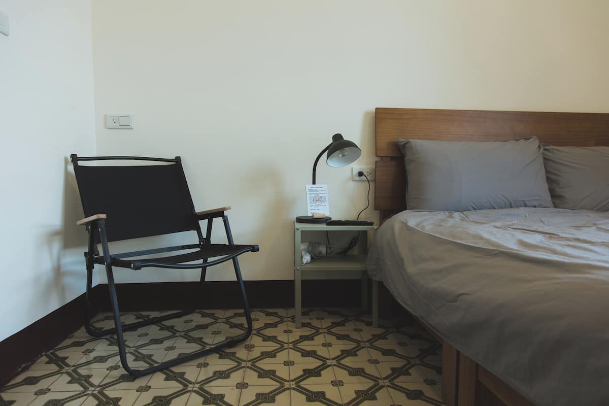 Quarto pequeno e bem organizado, com uma cama, uma cadeira, uma mesa com abajur e piso estampado.