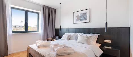 Quarto de hotel com cama grande, mesa de cabeceira, criado-mudo, quadro emoldurado na parede e janela com cortinas.