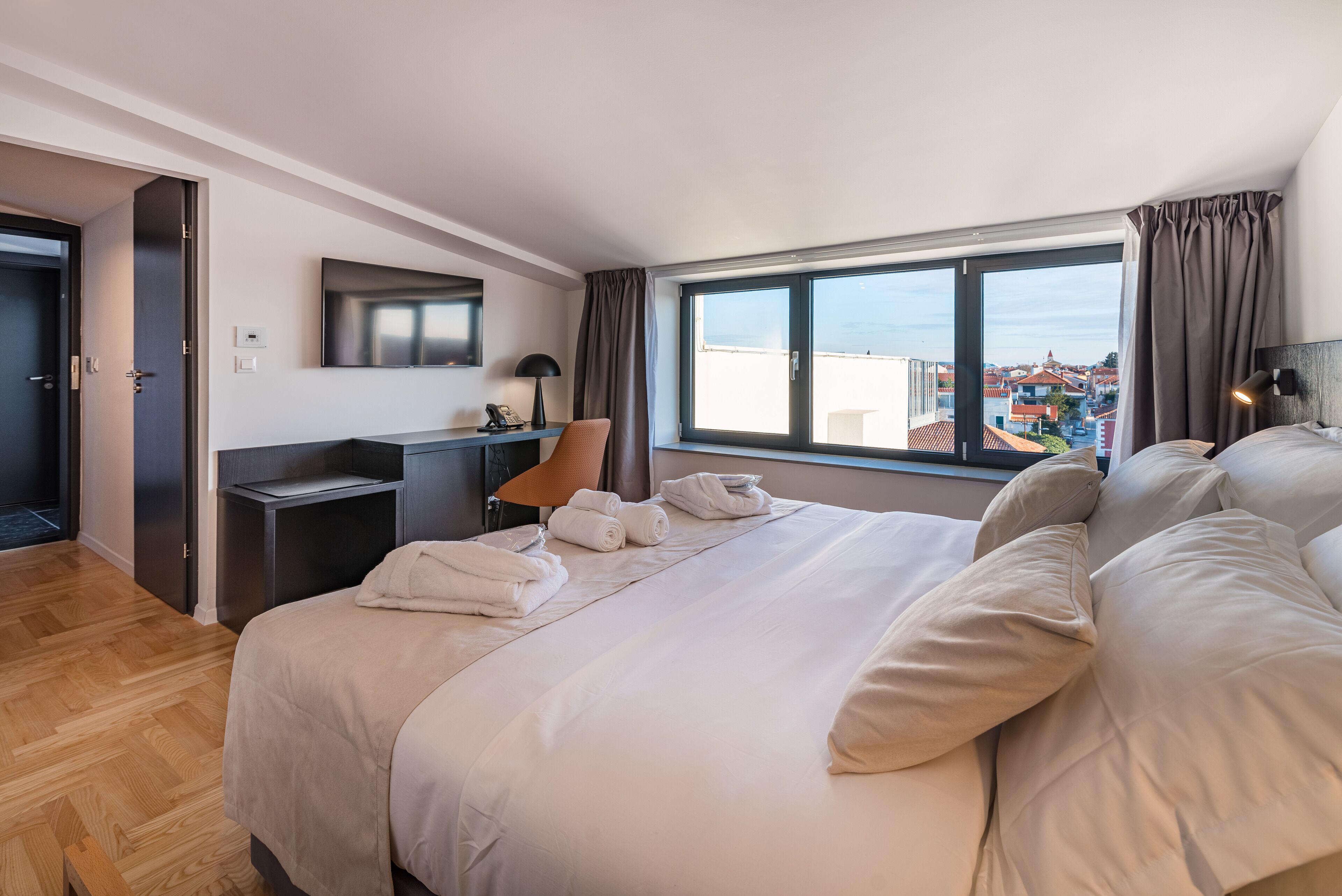 Quarto de hotel moderno com uma cama grande, uma escrivaninha, uma cadeira e vista para a cidade pela janela.