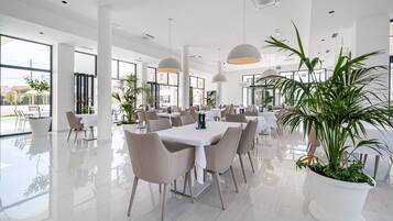 Uma sala de jantar espaçosa e moderna, com mesas e cadeiras brancas, janelas amplas e plantas em vasos.