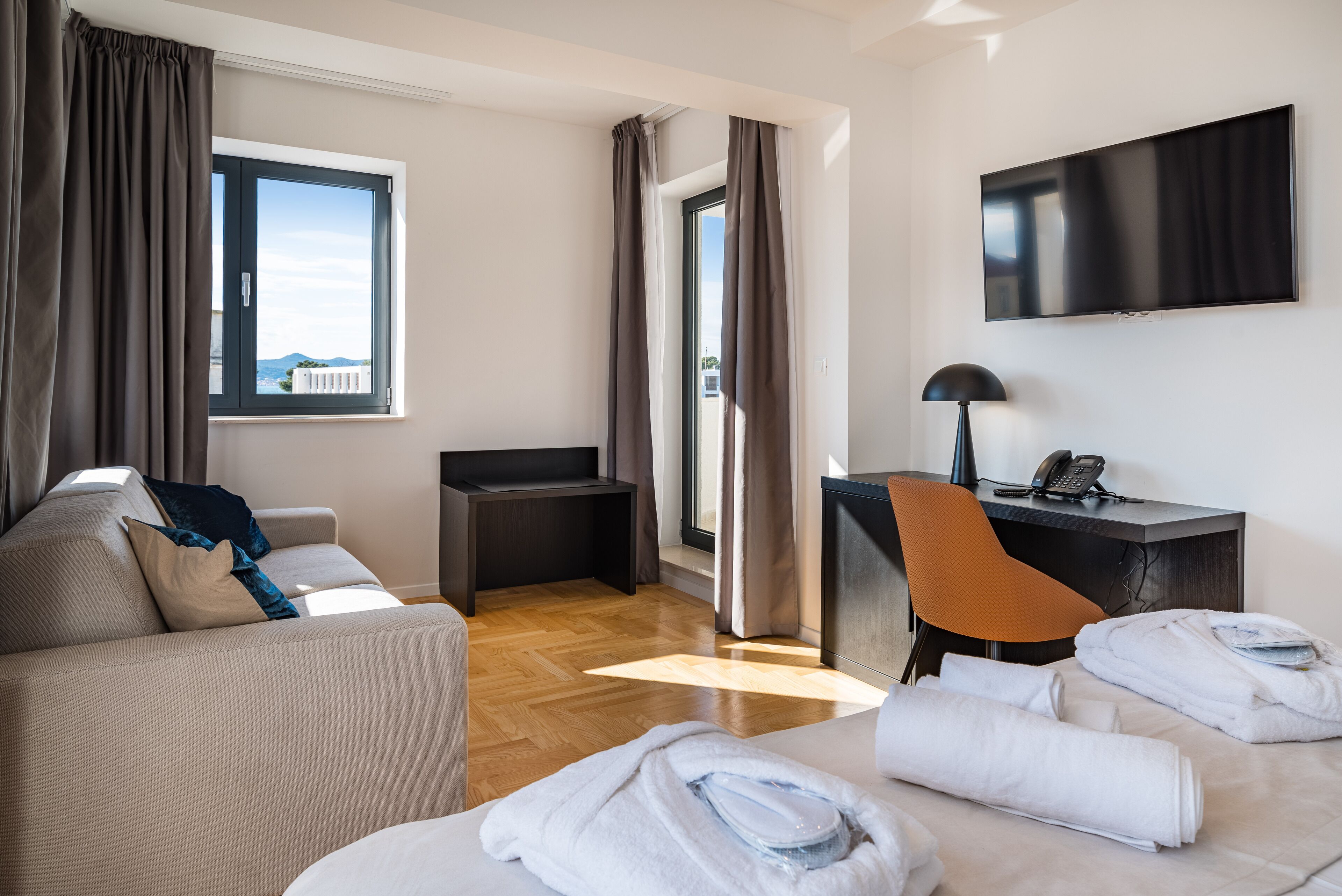 Quarto de hotel com sofá, escrivaninha, cadeira, TV e vista para o exterior.