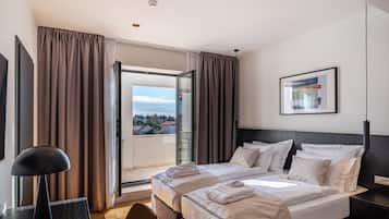Quarto de hotel com duas camas, uma janela grande com cortinas, uma escrivaninha com luminária e um quadro na parede.