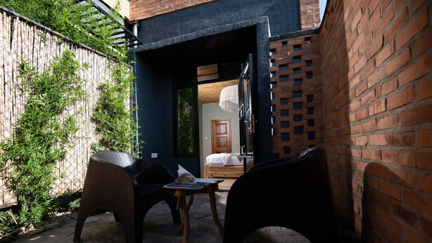 Uma área externa com assentos de vime, cadeiras e uma mesa, uma parede de tijolos e uma porta de madeira que dá para um quarto.