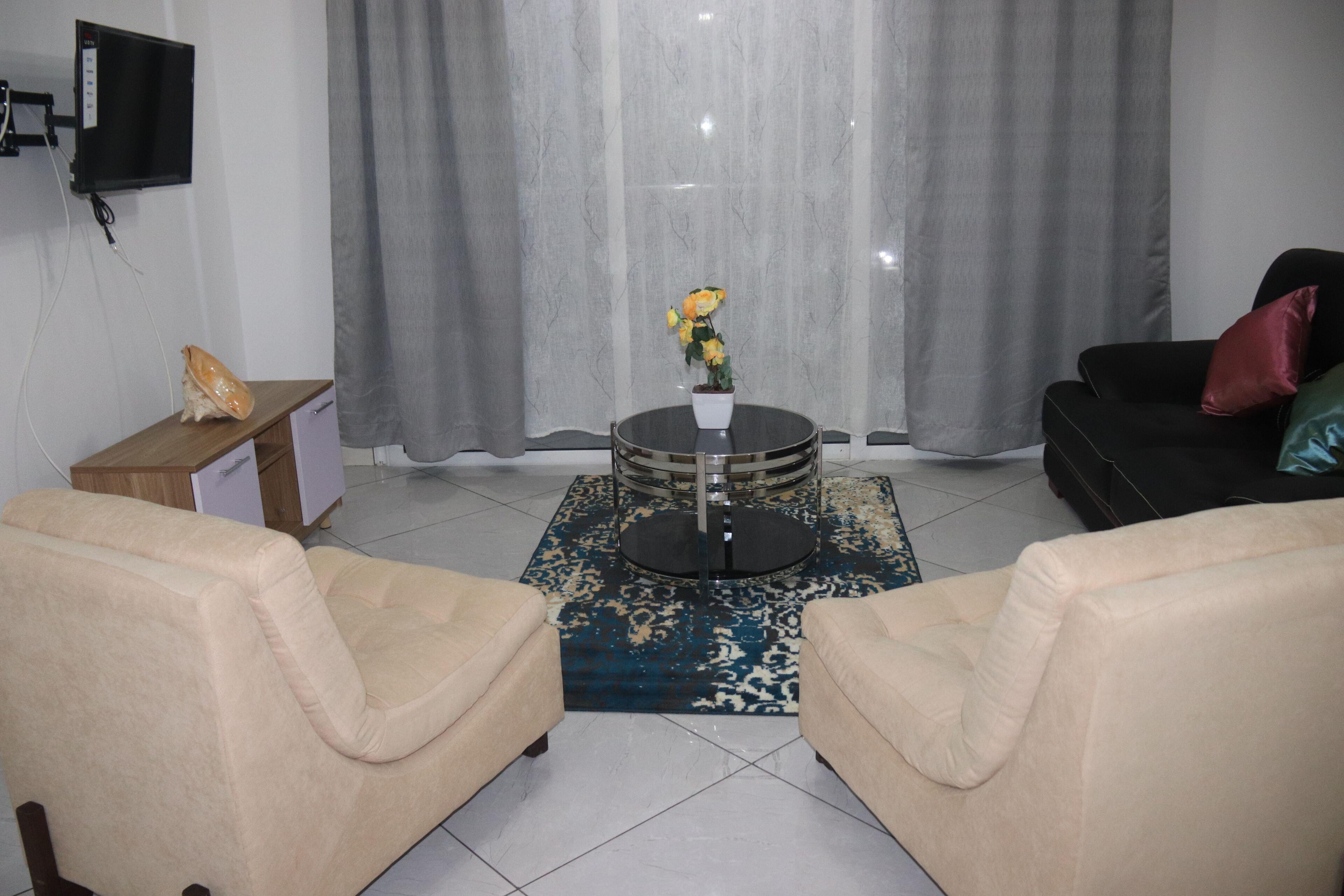 Uma sala de estar com dois poltronas bege, uma mesa de centro preta, uma pequena mesa lateral de madeira, uma TV de tela plana na parede e uma janela com cortinas cinzentas.