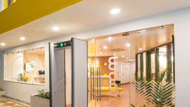 Entrada de hotel com balcão de recepção, área de atendimento e sala de espera com um sofá amarelo.