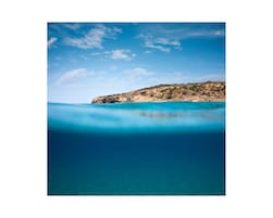 Paisagem costeira com águas azul-claras e uma colina rochosa ao fundo.