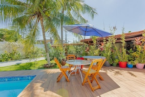 Uma área à beira da piscina com um deck de madeira, uma mesa redonda com cadeiras e um guarda-sol azul.