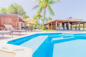 Piscina com espreguiçadeiras e guarda-sóis, cercada por estruturas de madeira e palmeiras.