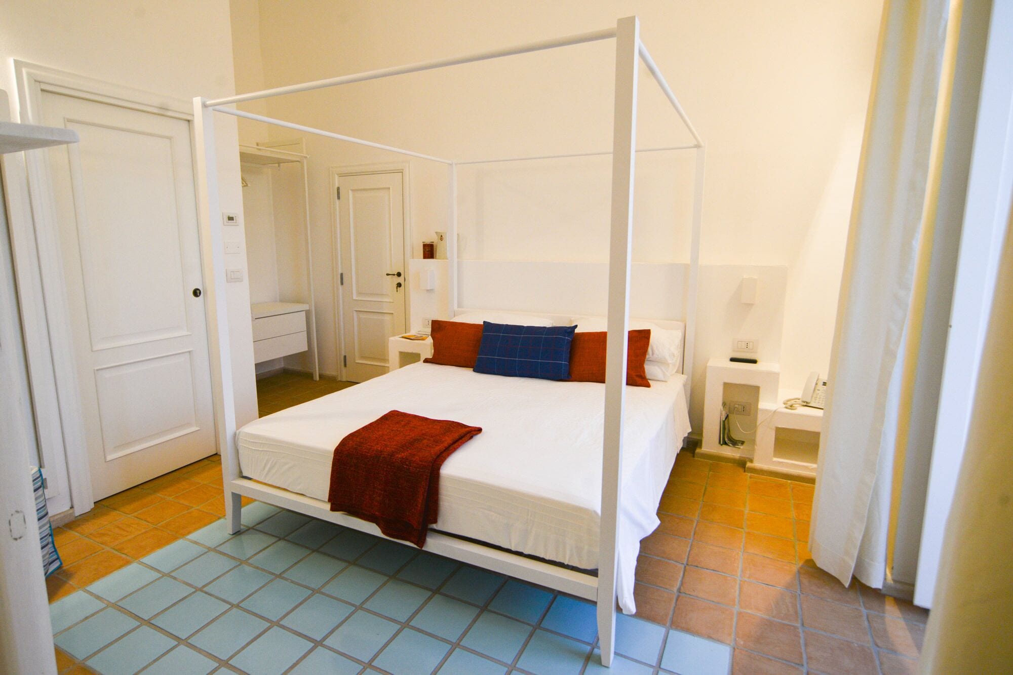 Uma cama de dossel com um cobertor vermelho, roupa de cama branca e duas almofadas. Há uma mesa de cabeceira com uma luminária e um telefone. O quarto tem paredes brancas e piso de azulejos.