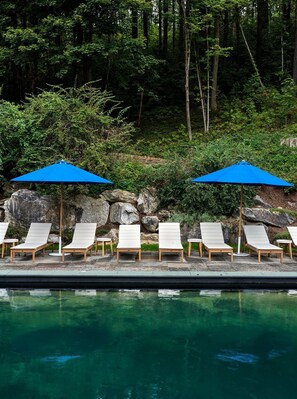 Área da piscina com espreguiçadeiras e guarda-sóis azuis, emoldurada por uma paisagem rochosa e vegetação densa.