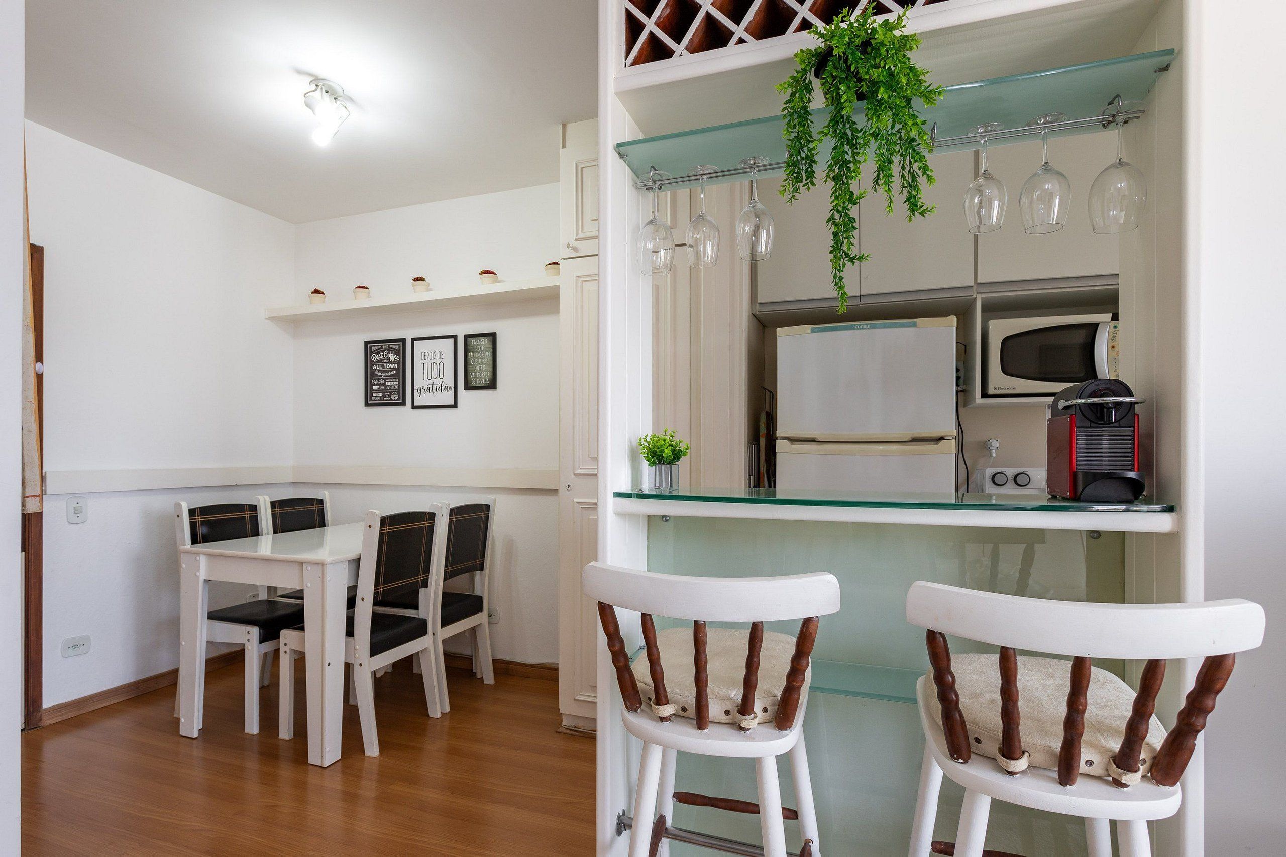 Espaço de refeições com mesa e cadeiras brancas, balcão de bar com tampo de vidro e planta pendente, e um micro-ondas acima.