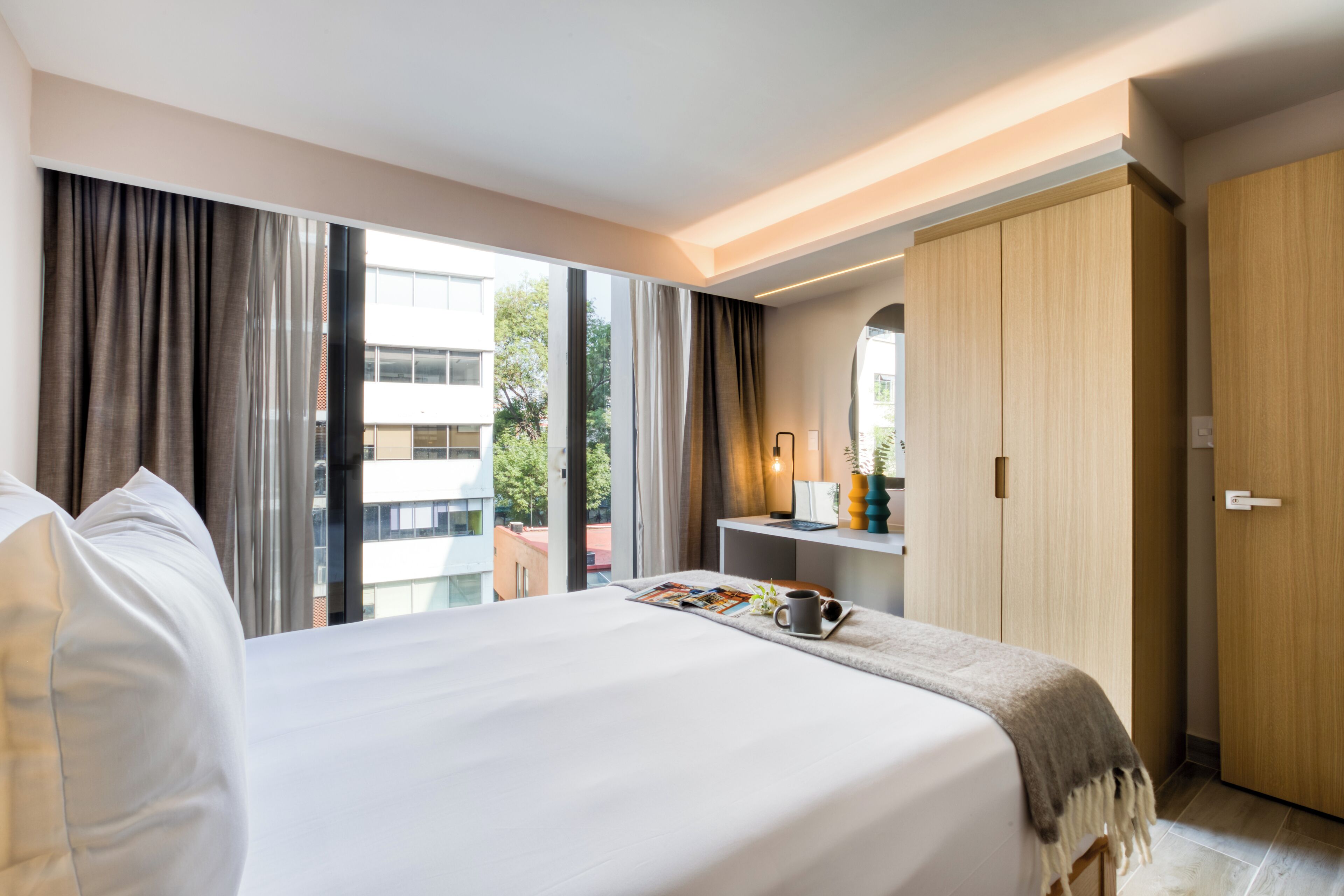 Quarto de hotel com uma cama grande, uma escrivaninha com abajur, um guarda-roupa e vista para edifícios.