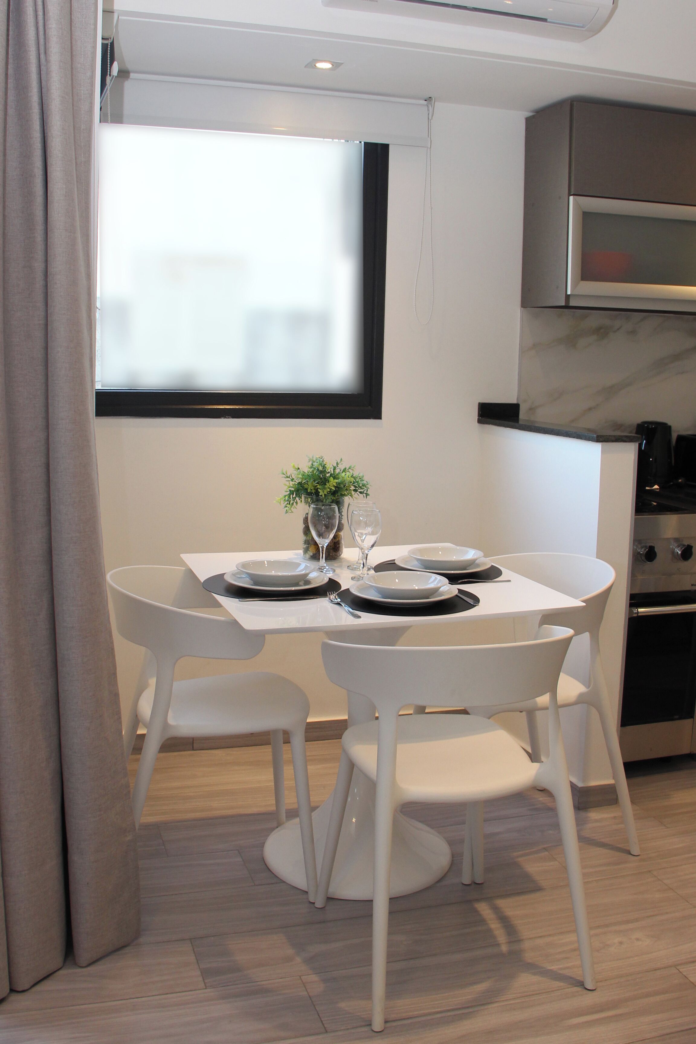 Uma sala de jantar moderna com uma mesa posta para quatro, cadeiras brancas, um pequeno vaso de planta e uma janela com cortinas.