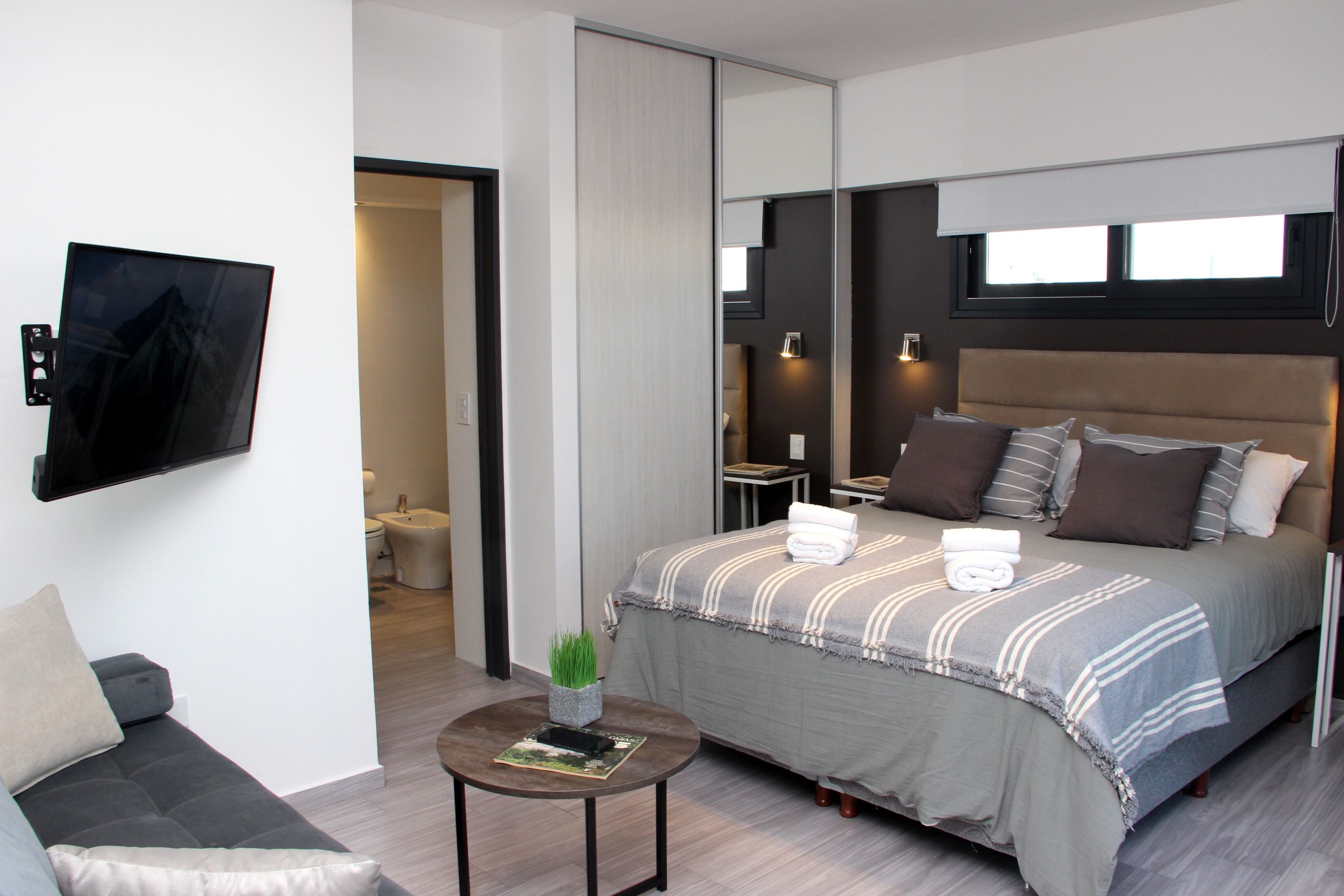 Quarto moderno com uma cama grande, uma mesinha redonda com uma planta, um sofá e uma televisão fixada na parede.